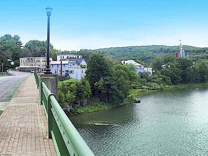 The green bridge spans calm waters leading to white buildings nestled among trees, creating a scene of tranquil riverside beauty.