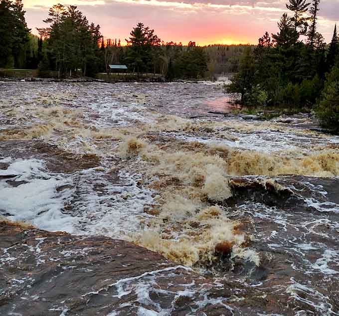The sky turns a beautiful shade of pink as the sun sets behind the pines, casting shadows across the river.