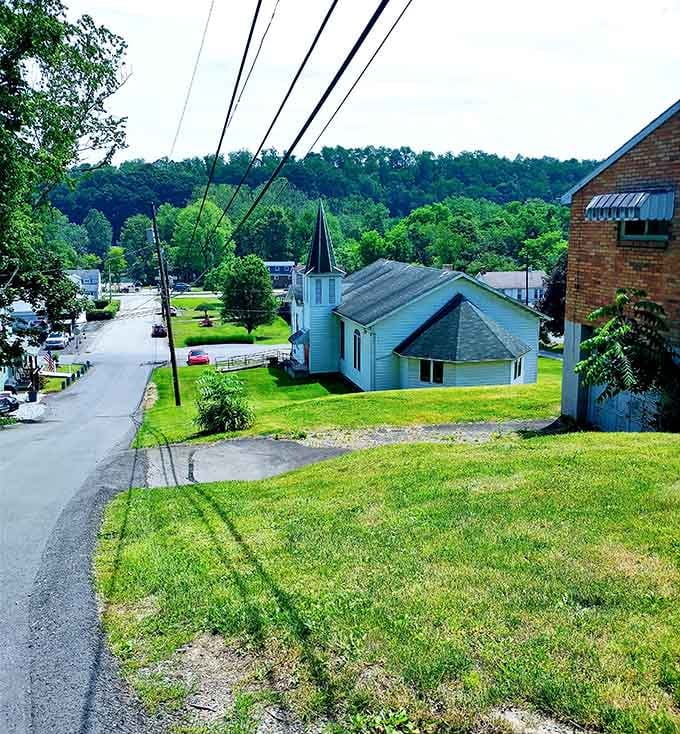Look down the grassy hillside at a lovely white church nestled beautifully within a peaceful, tree-lined mountain valley community.