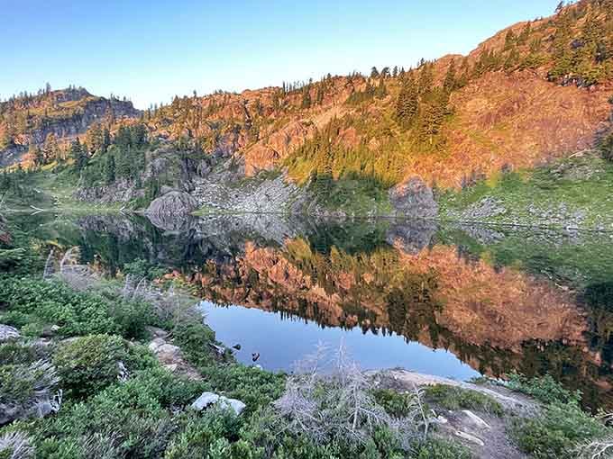 Golden hour light painting the cliffs and trees in warm copper tones, reflected perfectly in the glassy, still water below.