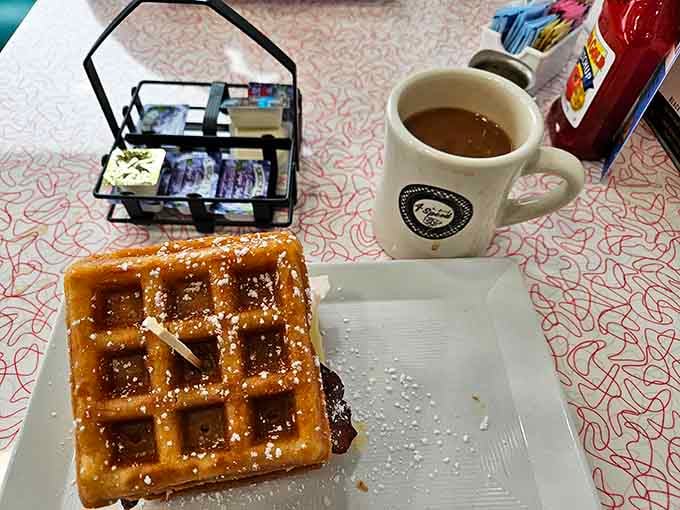 A golden waffle dusted with powdered sugar, paired with a steaming branded mug. Breakfast just got its act together.