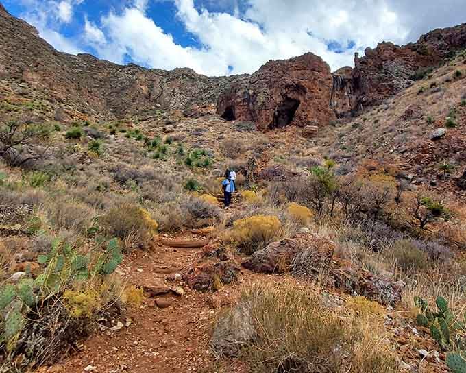 Two hikers climb toward dark cave openings in rust-red Franklin Mountains rock, chasing views that reward every breathless step.