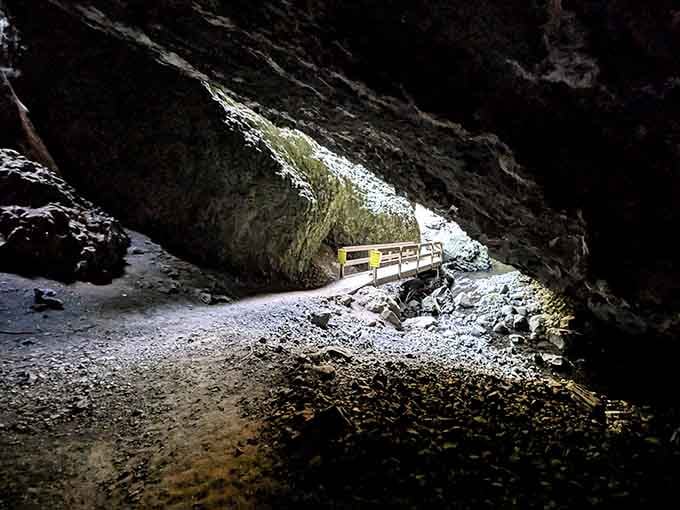 A glowing arch of daylight at the cave entrance frames a wooden bridge, like nature designed its own dramatic stage exit.