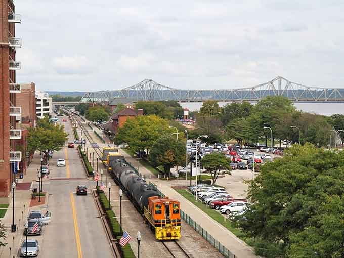 A bright orange locomotive rolls along Peoria's riverfront boulevard, with a graceful bridge stretching wide across the Illinois River beyond.