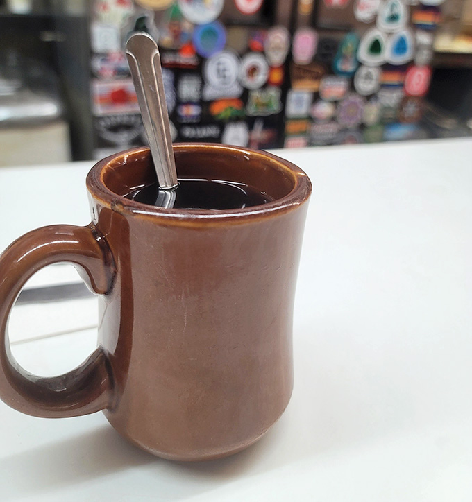 That sturdy brown mug filled with dark, steaming coffee, with a wall of stickers blurred behind it, is pure diner poetry.