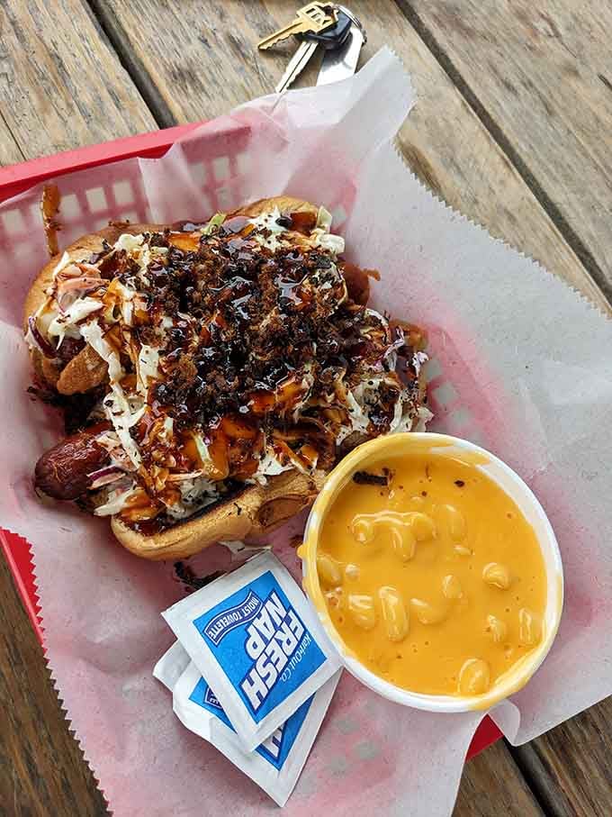 A loaded all-beef dog buried under coleslaw, crispy bits, and BBQ drizzle, with cheesy mac alongside for moral support.
