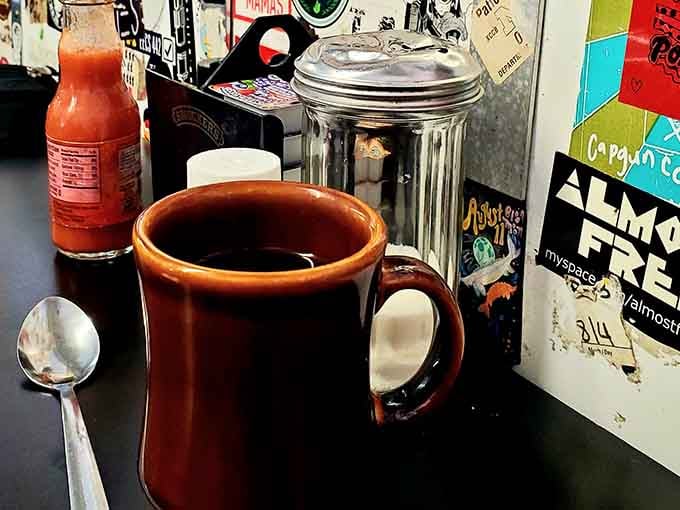 A sturdy diner mug filled with hot coffee, surrounded by stickers and hot sauce. The most honest cup in Ann Arbor.