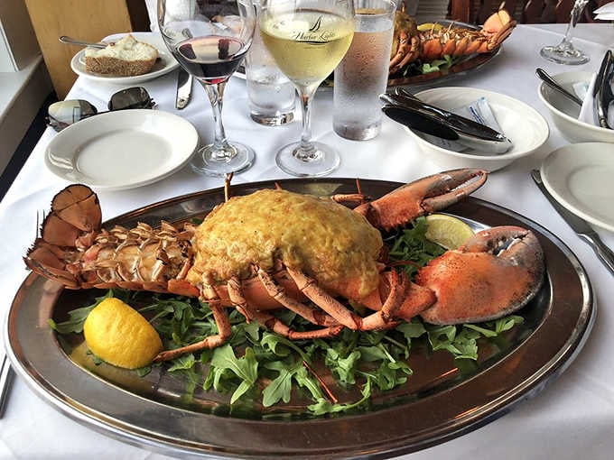 A whole stuffed lobster on a silver platter, arugula beneath, wine glasses in the background. This is a full production.