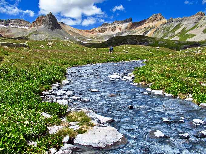 A crystal-clear mountain stream tumbles over white rocks through a lush alpine meadow, cheerfully leading hikers toward the basin above.