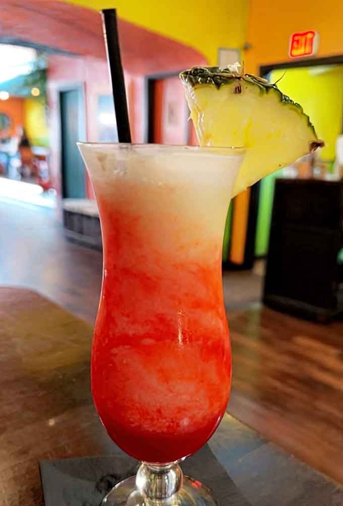 A layered tropical cocktail with a pineapple wedge perched on top, served against a backdrop of pure Caribbean color.