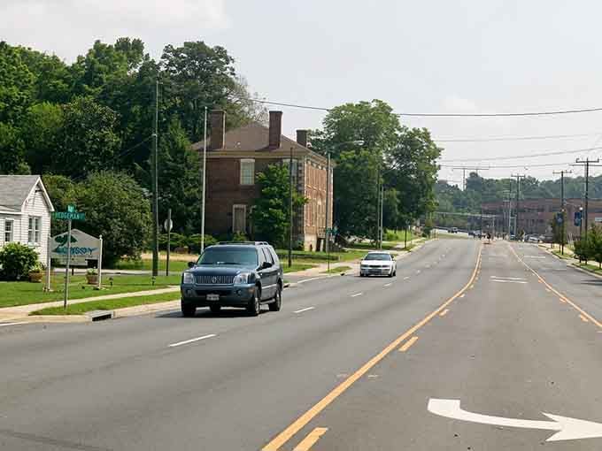 Cars travel along an open roadway featuring a historic two-story brick house surrounded by lush, mature green trees.