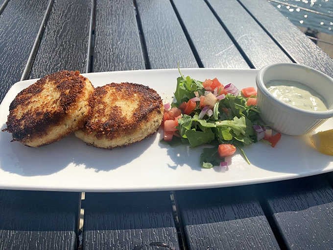 Two golden crab cakes beside a bright tomato salad and creamy dipping sauce, with the harbor sparkling just beyond.