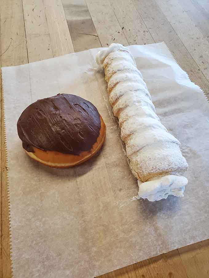 A chocolate-glazed donut and a cream horn sharing parchment paper like old friends. Pick one. Actually, don't. Get both.