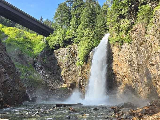 The bridge overhead frames this dramatic drop where water thunders into a turquoise pool with raw mountain energy.