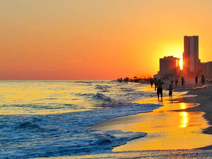 Golden light pours across the shoreline as beachgoers stroll through the warm surf of a Gulf Shores evening.